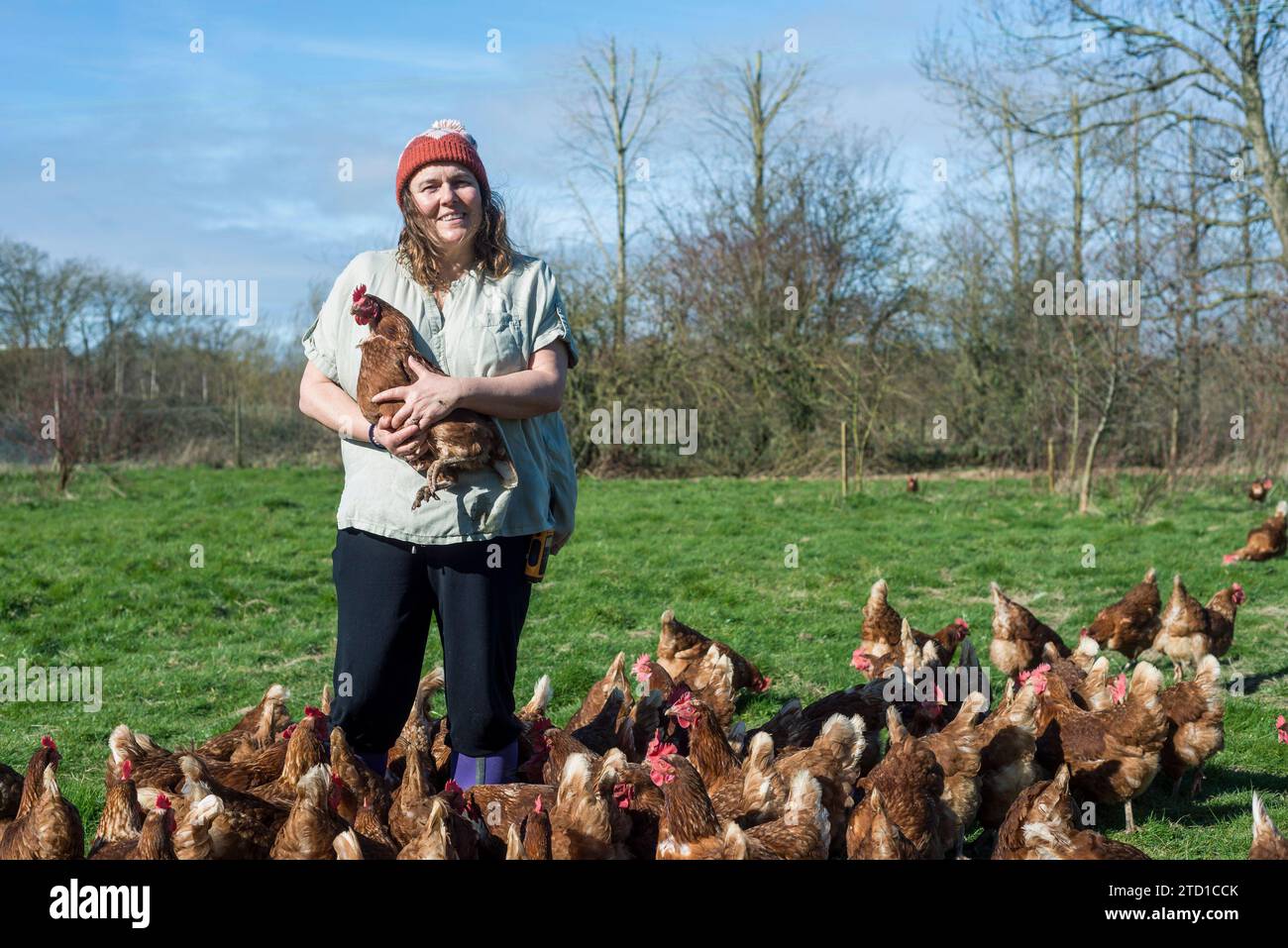 A female farmer stands holding a chicken surrounded by a flock of ...
