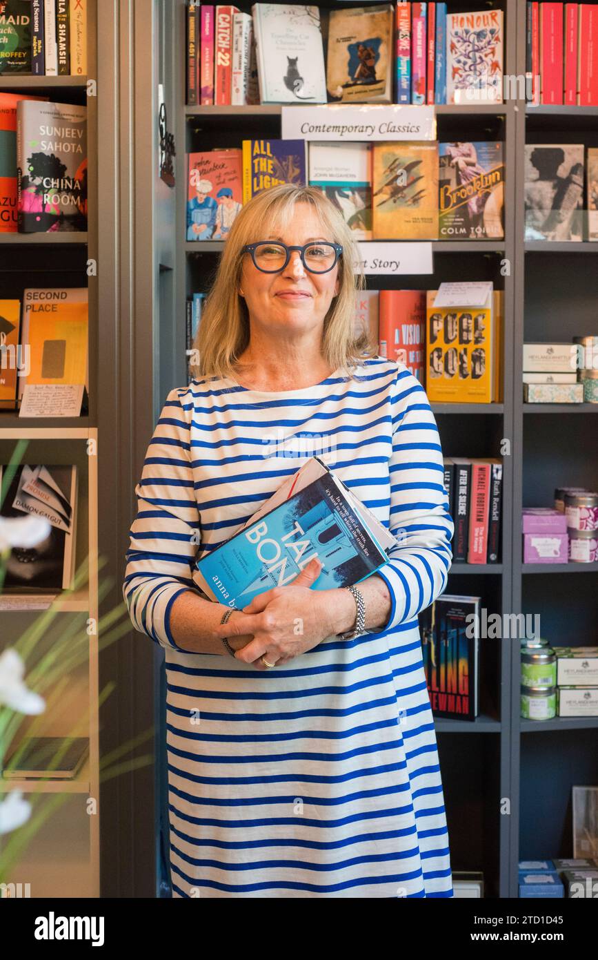 A happy book seller stands in her shop holding a few books in front of ...