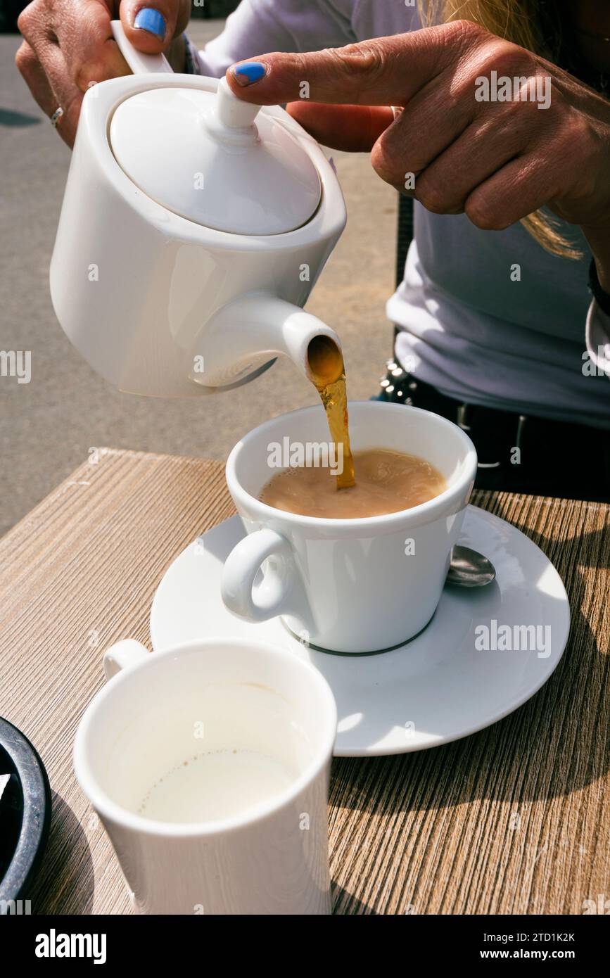 A person pours a cup of tea from a white teapot into a white cup and ...