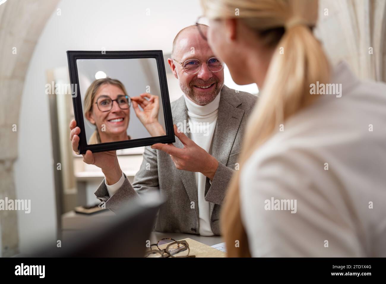 Optician holding a mirror to smiling woman trying on new glasses in a ...