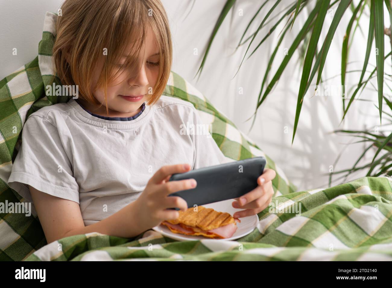 A young boy lies in bed looking at his phone and eating a sandwich ...