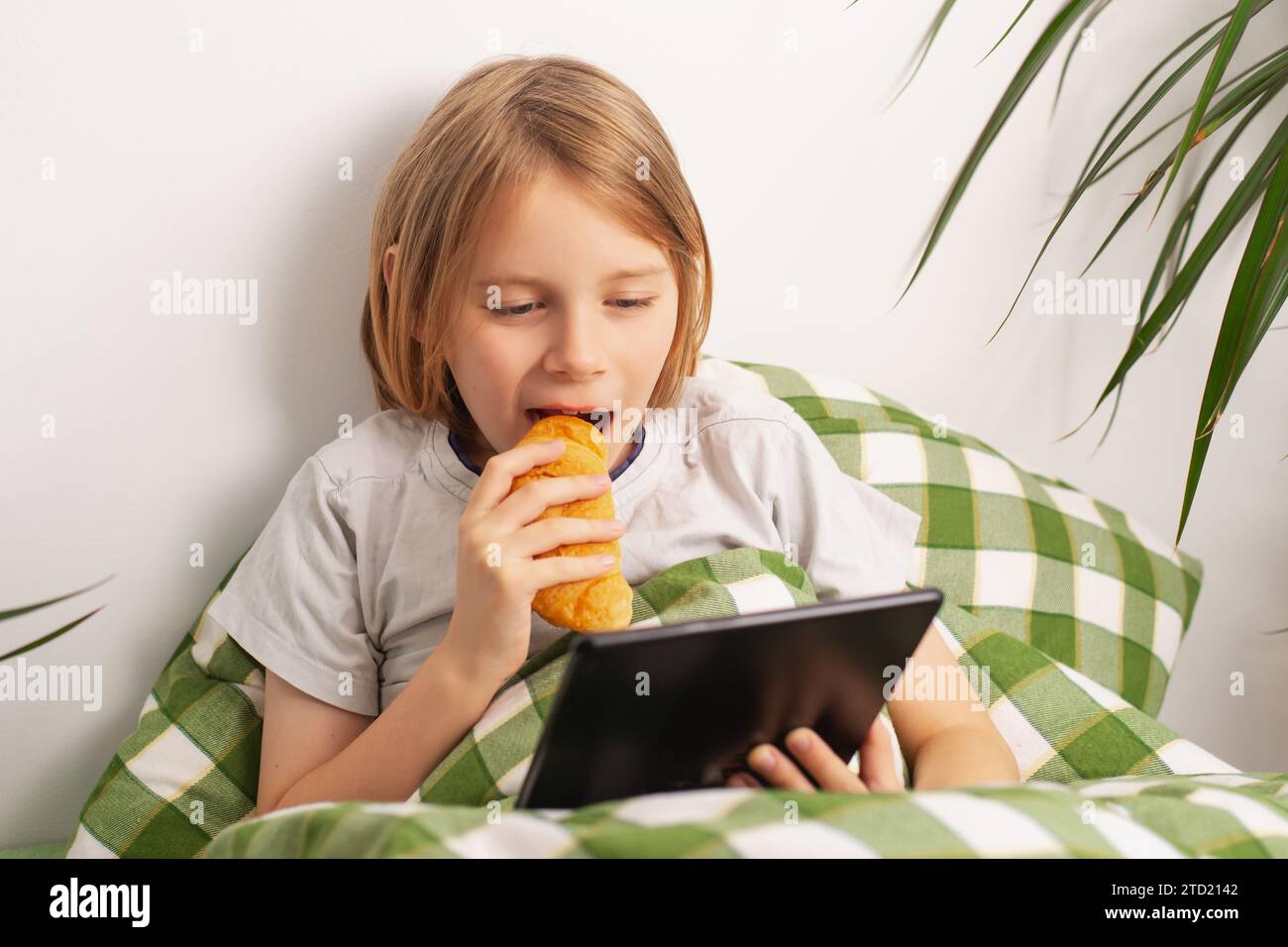 A young boy lounges on the bed, engrossed in his tablet, snacking on a ...