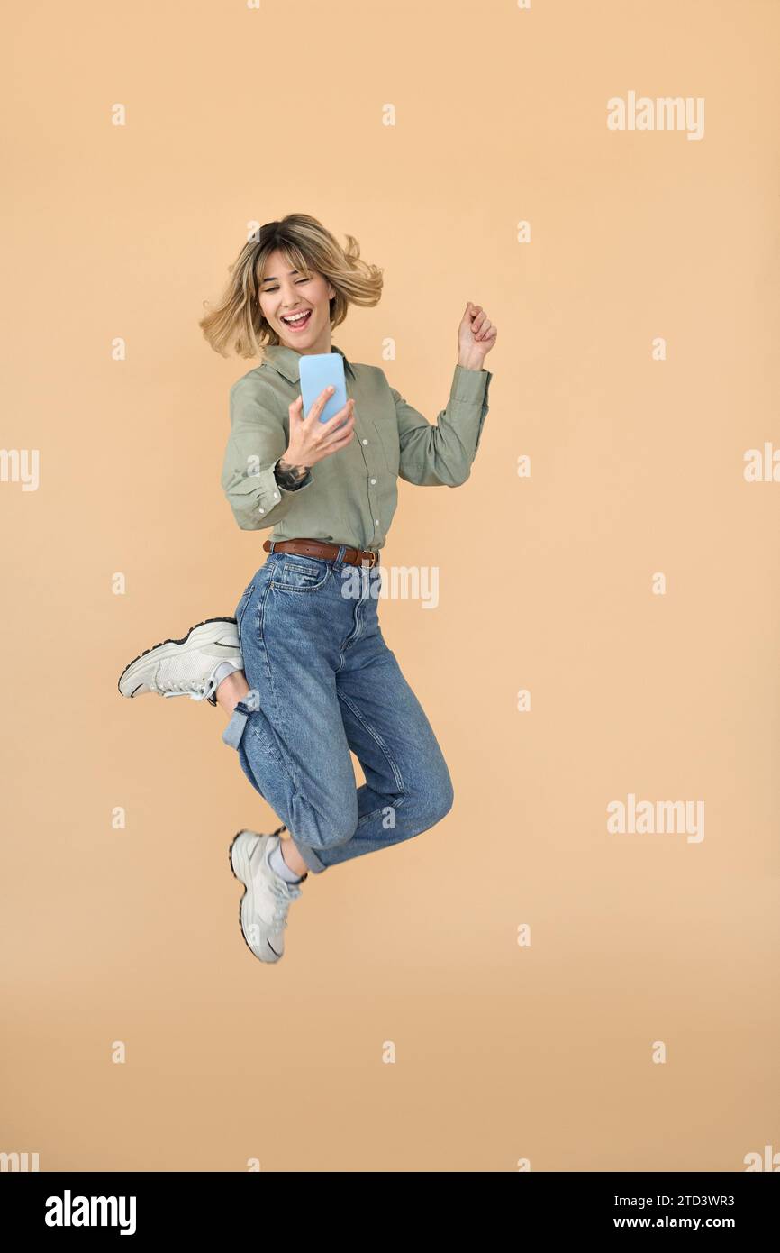 Happy gen z blonde young woman using smartphone jumping isolated on ...
