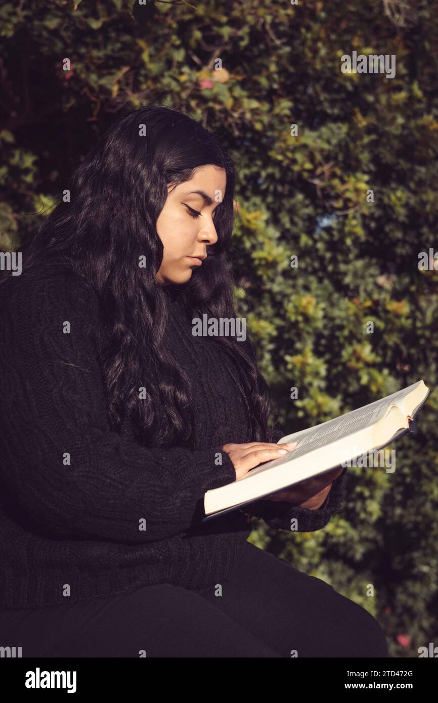 Young woman, seated attentively reading a book in the garden vertical ...