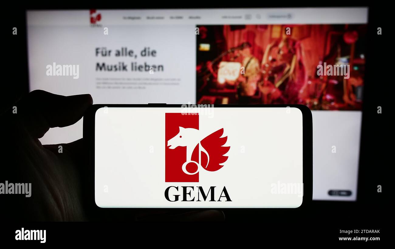 Person holding mobile phone with logo of German collecting society GEMA in front of web page ...