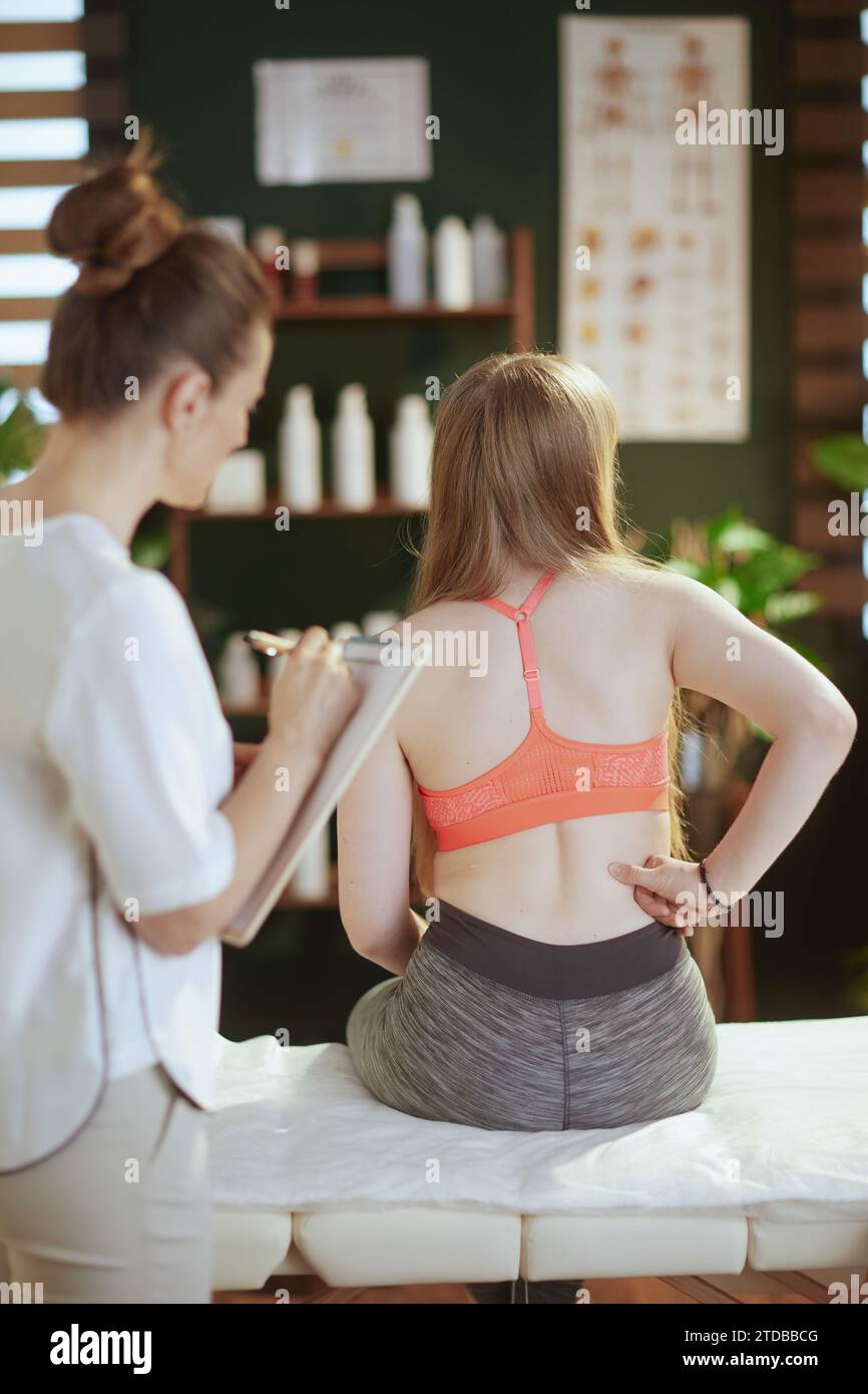 Healthcare time. Seen from behind female massage therapist in massage cabinet with clipboard and ...