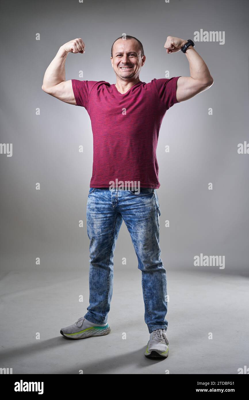 Middle aged man in tee and jeans flexing his biceps on gray background ...