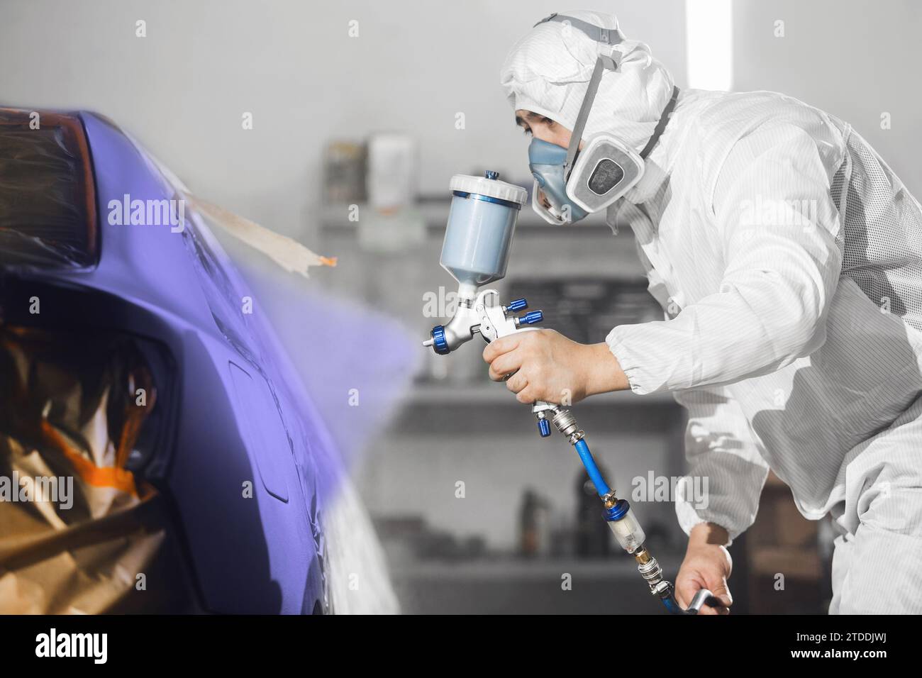 Male worker in protective clothes and mask painting car body to blue ...