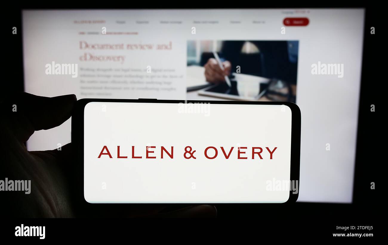 Person holding cellphone with logo of British law firm Allen and Overy LLP in front of business ...
