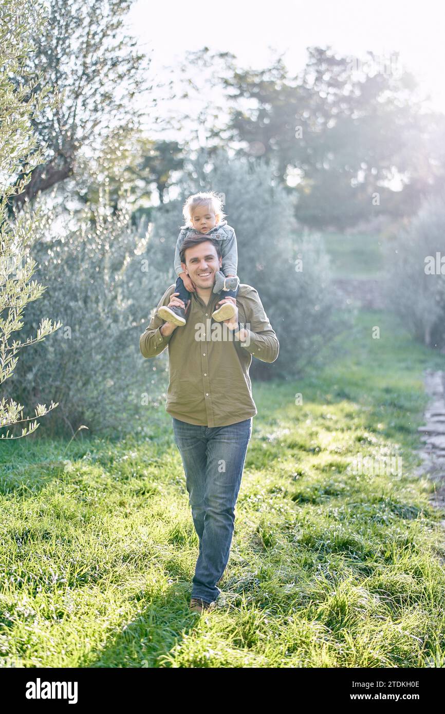 Smiling dad with a little girl on his shoulders walks through the green grass in the park Stock ...