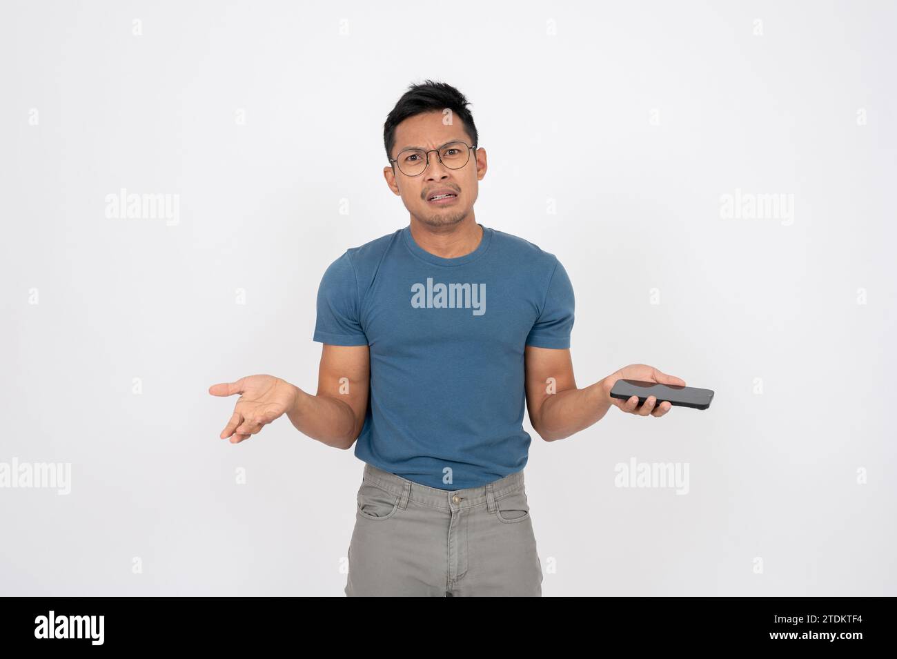 A doubtful, confused Asian man with a phone in his hand is shrugging ...