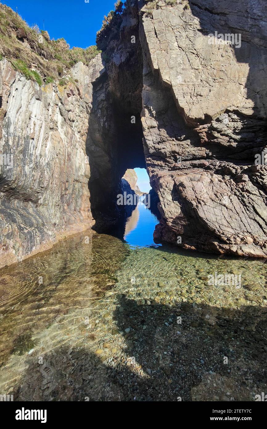 natural archway leads to a clear pool, reflecting the sky amidst rugged ...