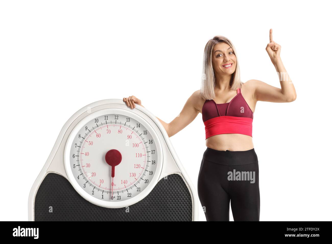 Young woman in sportswear leaning on a big weight scale and pointing up ...