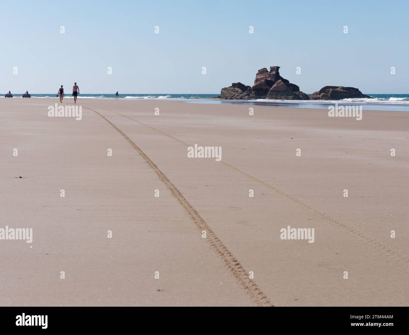 Couple people walk on a sandy beach with rock outcrop and quad bikes ...