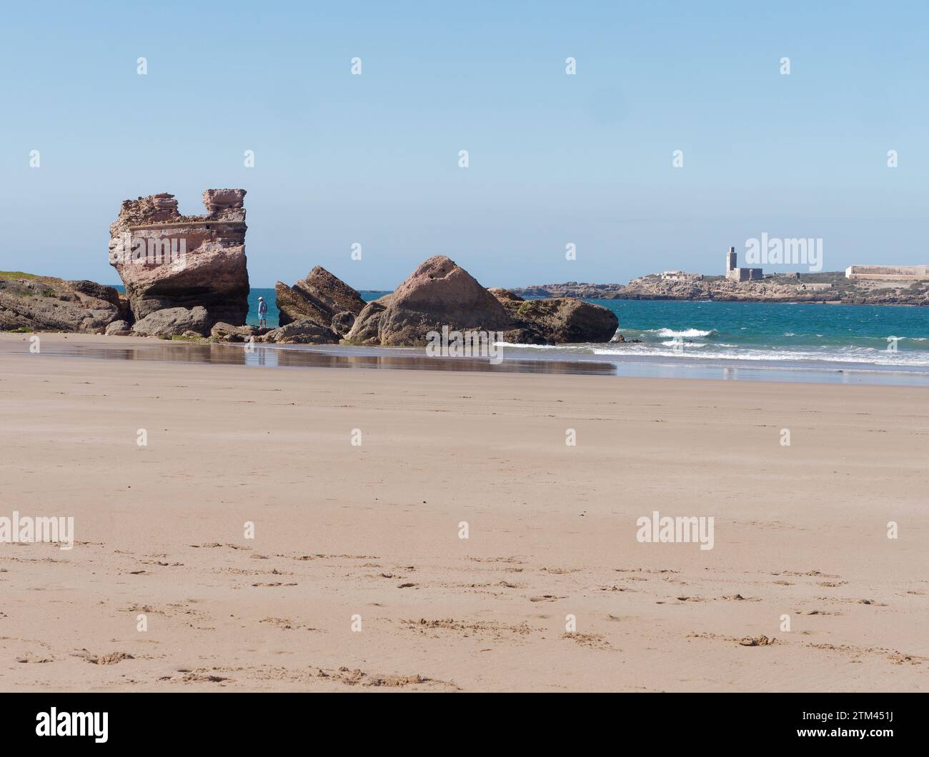 Man explores a rock outcrop off a sandy beach with an island behind in ...