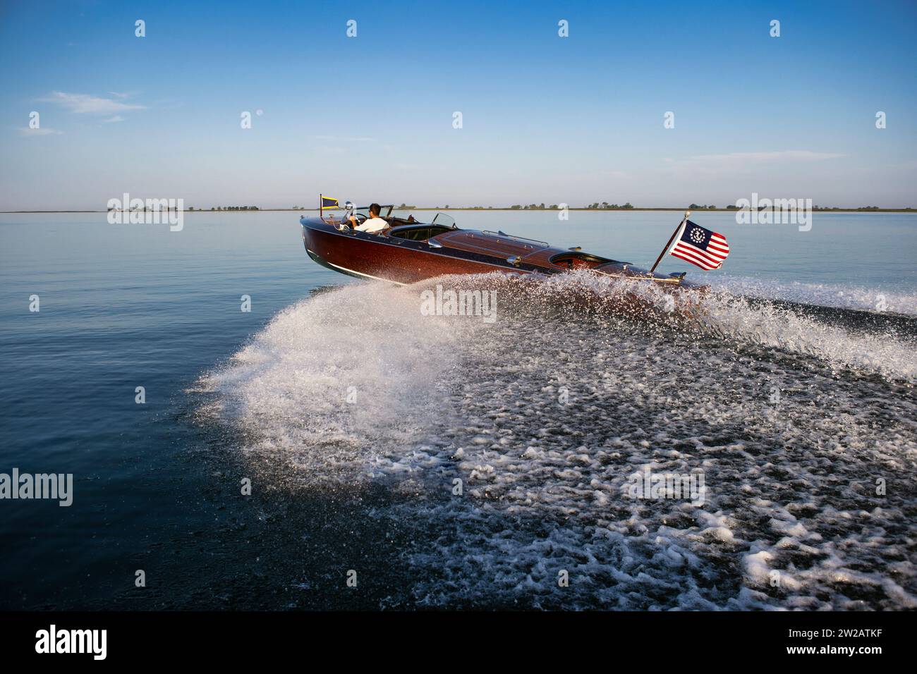 A wood, triple-cockpit speedboat throwing spray while crossing a calm ...