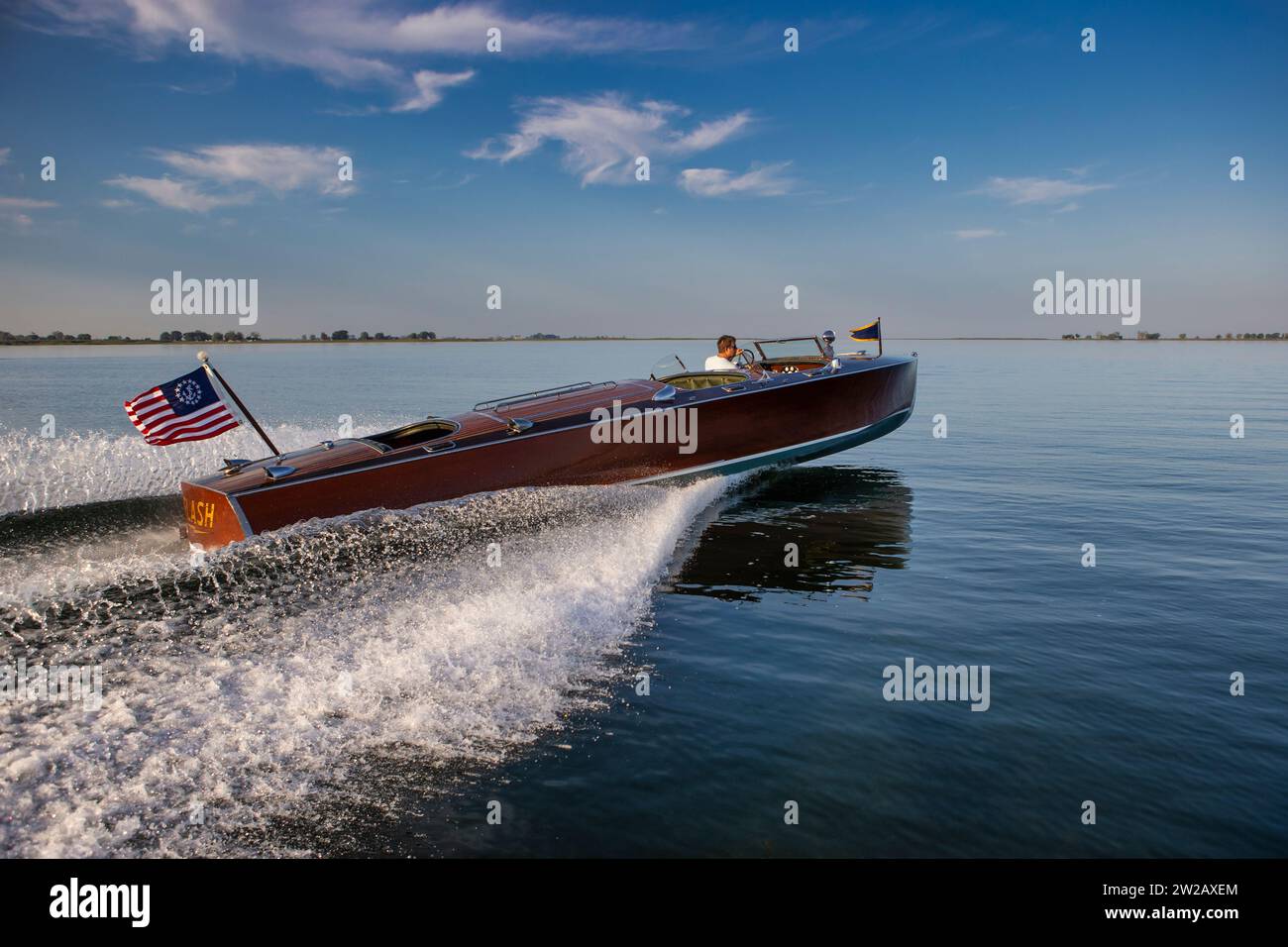 A triple-cockpit, wooden speedboat running at high speed on a calm lake ...