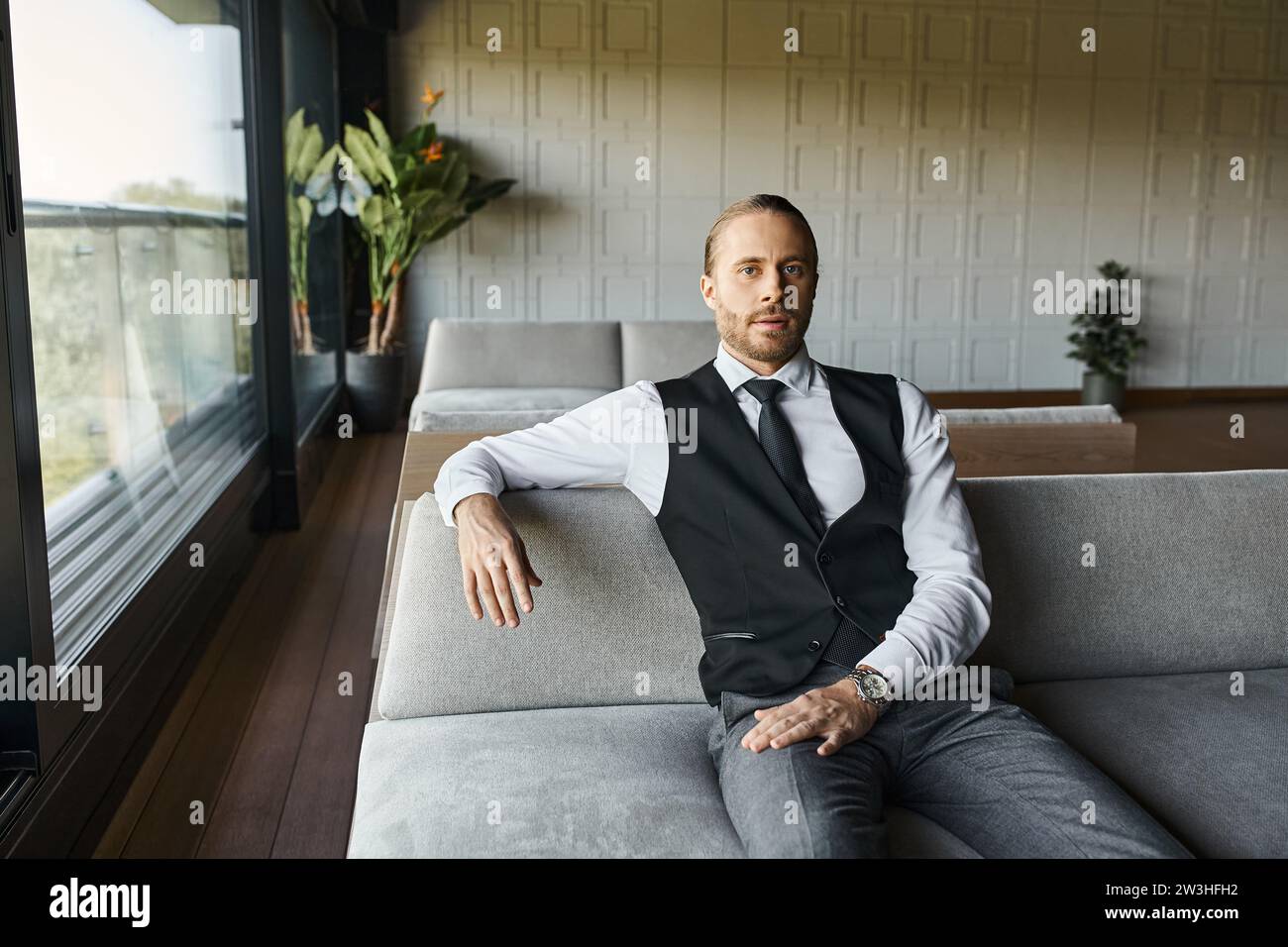 handsome elegant man in black smart vest sitting on sofa and looking at ...