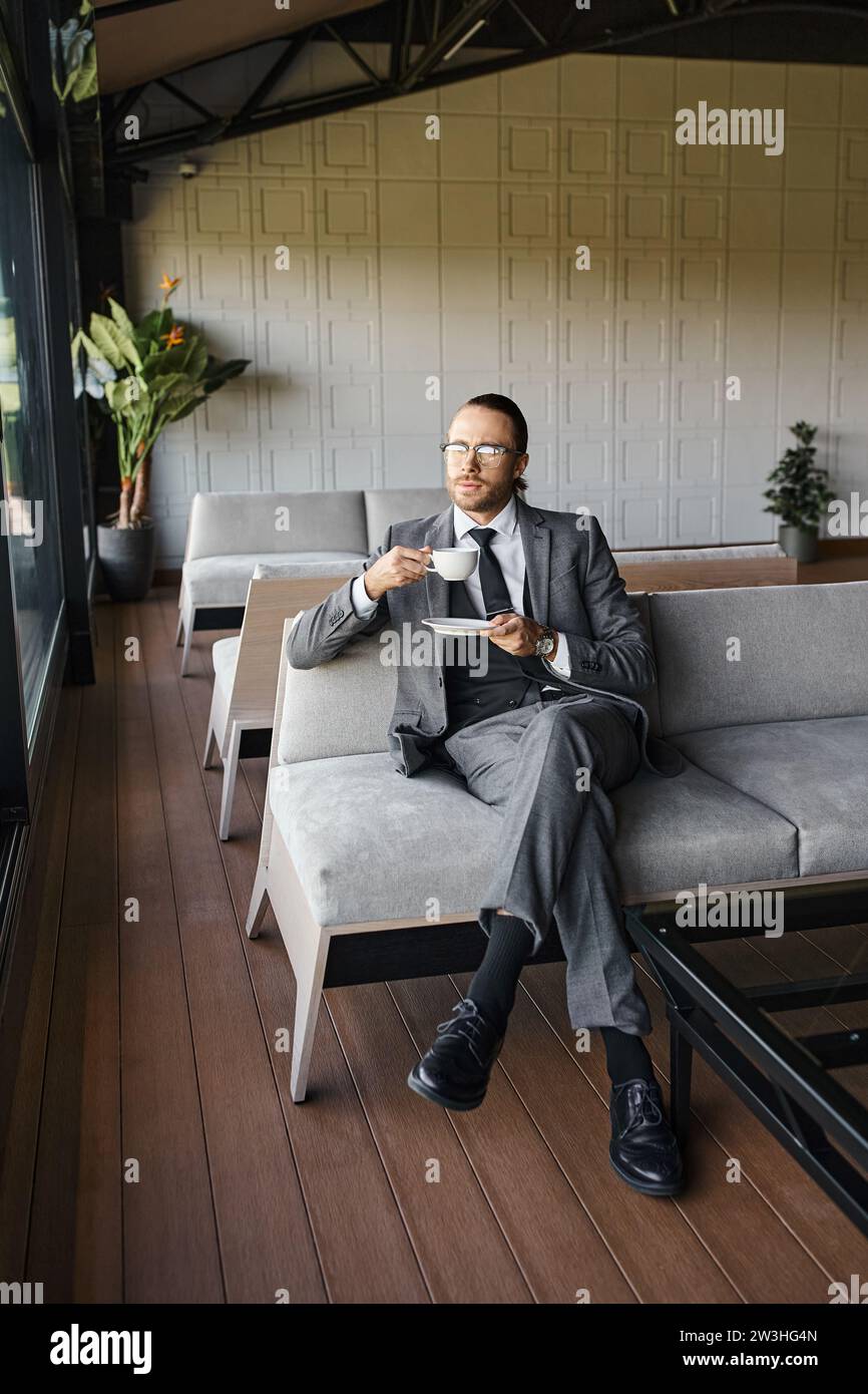 good looking elegant man in chic gray suit sitting on sofa and drinking ...