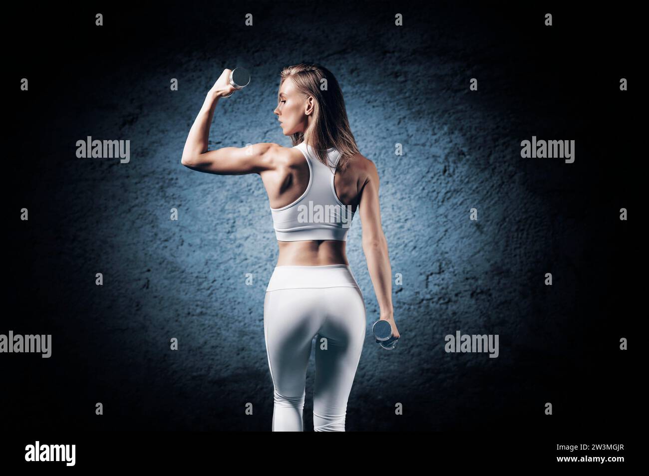 Adorable girl is training in the gym. She lifts the dumbbells up. Back ...