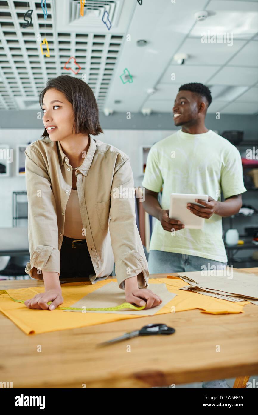 smiling asian designer measuring sewing pattern near african american ...