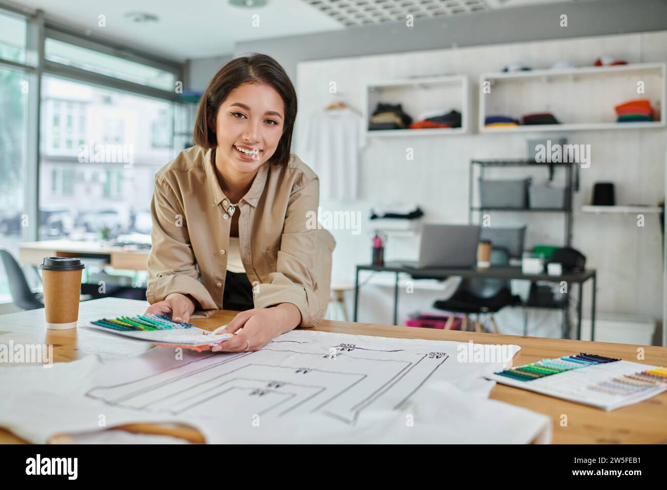 joyful creative asian designer with color samples near clothes with ...