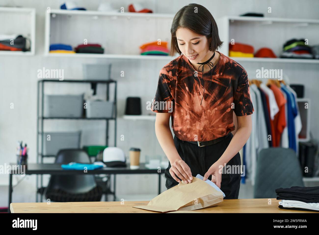 happy asian woman packing neatly folded garments on table in textile ...