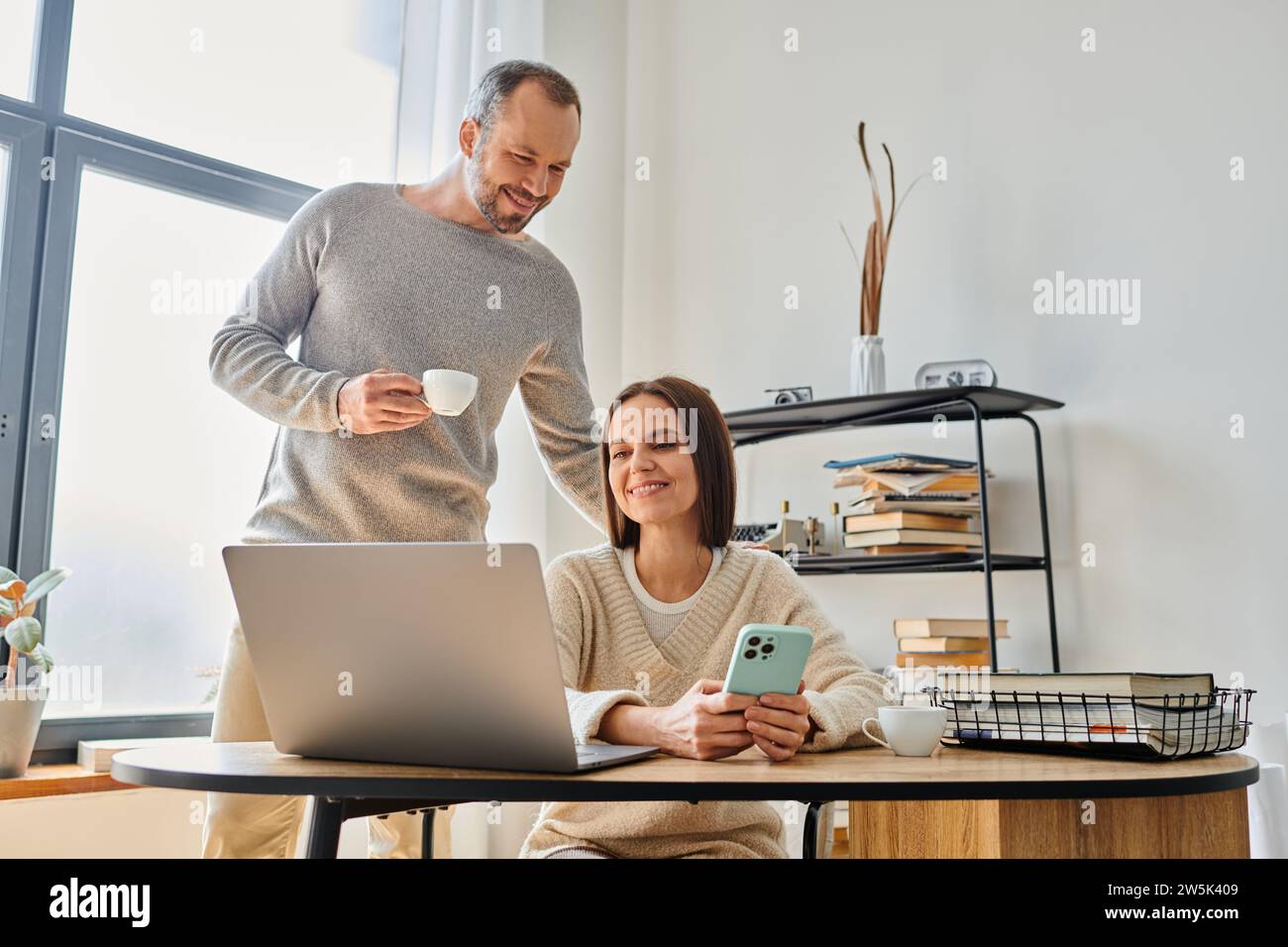 smiling man with coffee cup supporting wife working on laptop at home ...