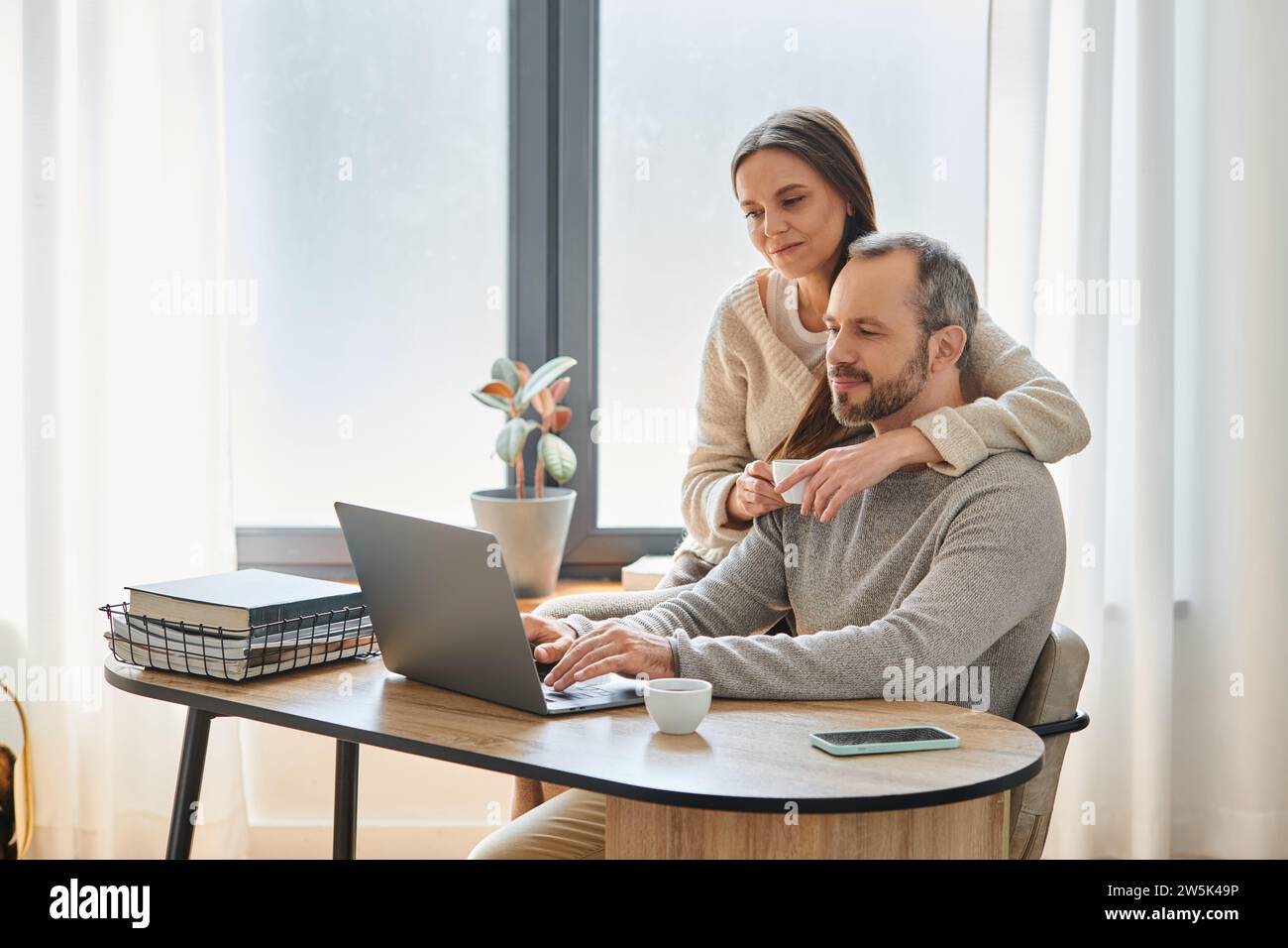 supportive wife with coffee cup hugging husband working on laptop in ...