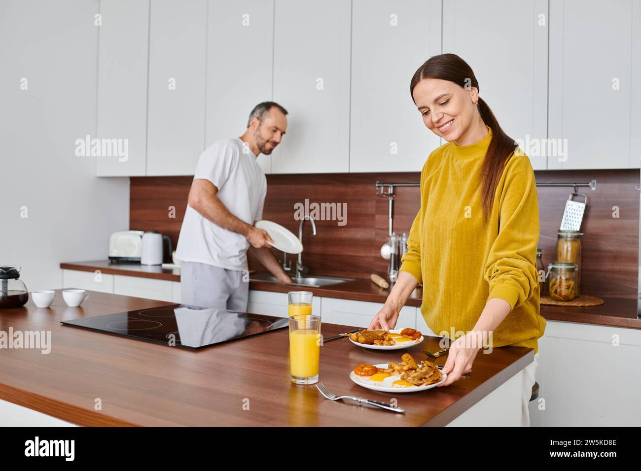smiling woman serving tasty breakfast while husband washing dishes in kitchen, care and harmony ...