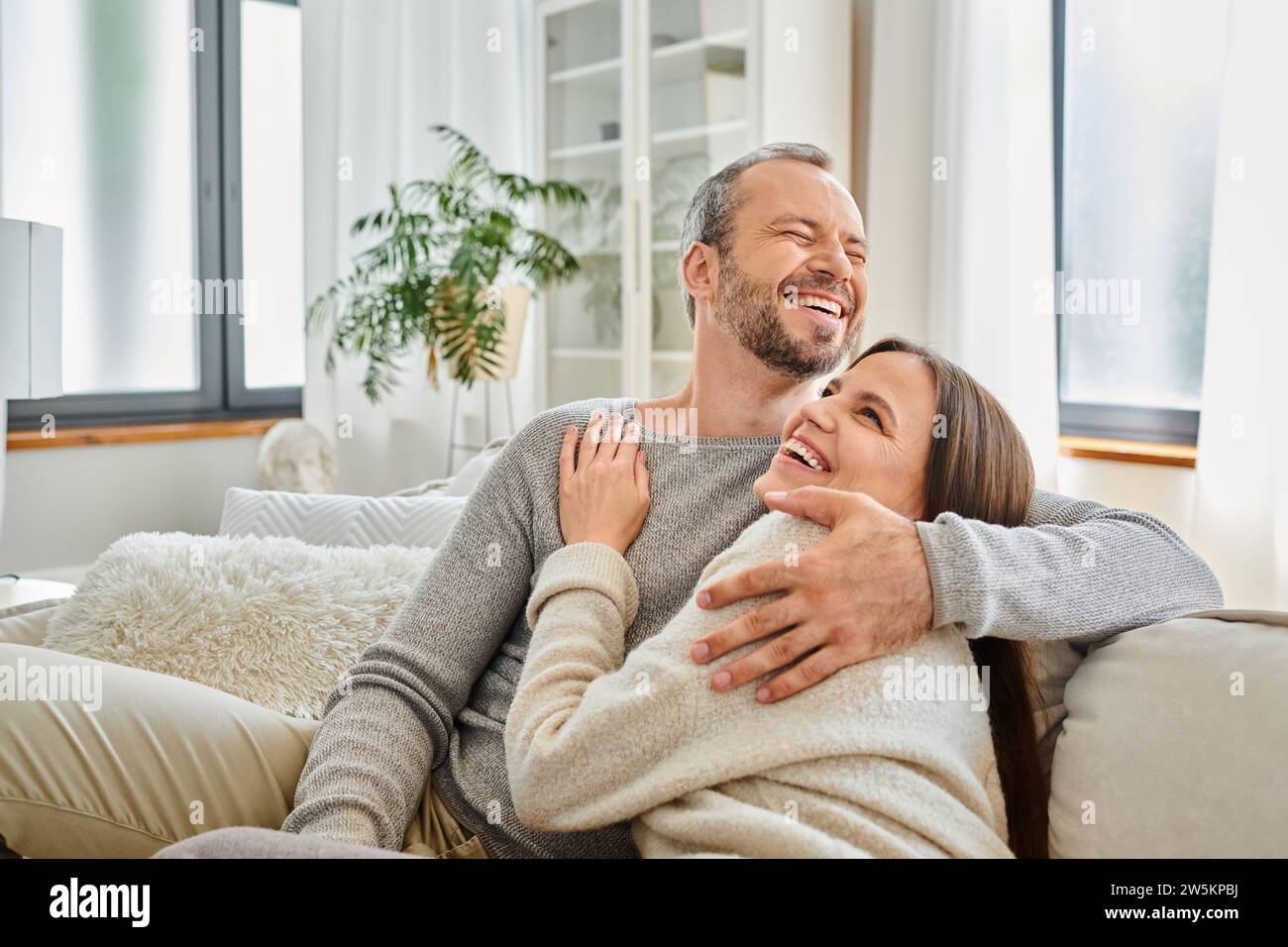 excited child-free couple embracing and laughing on couch in modern ...