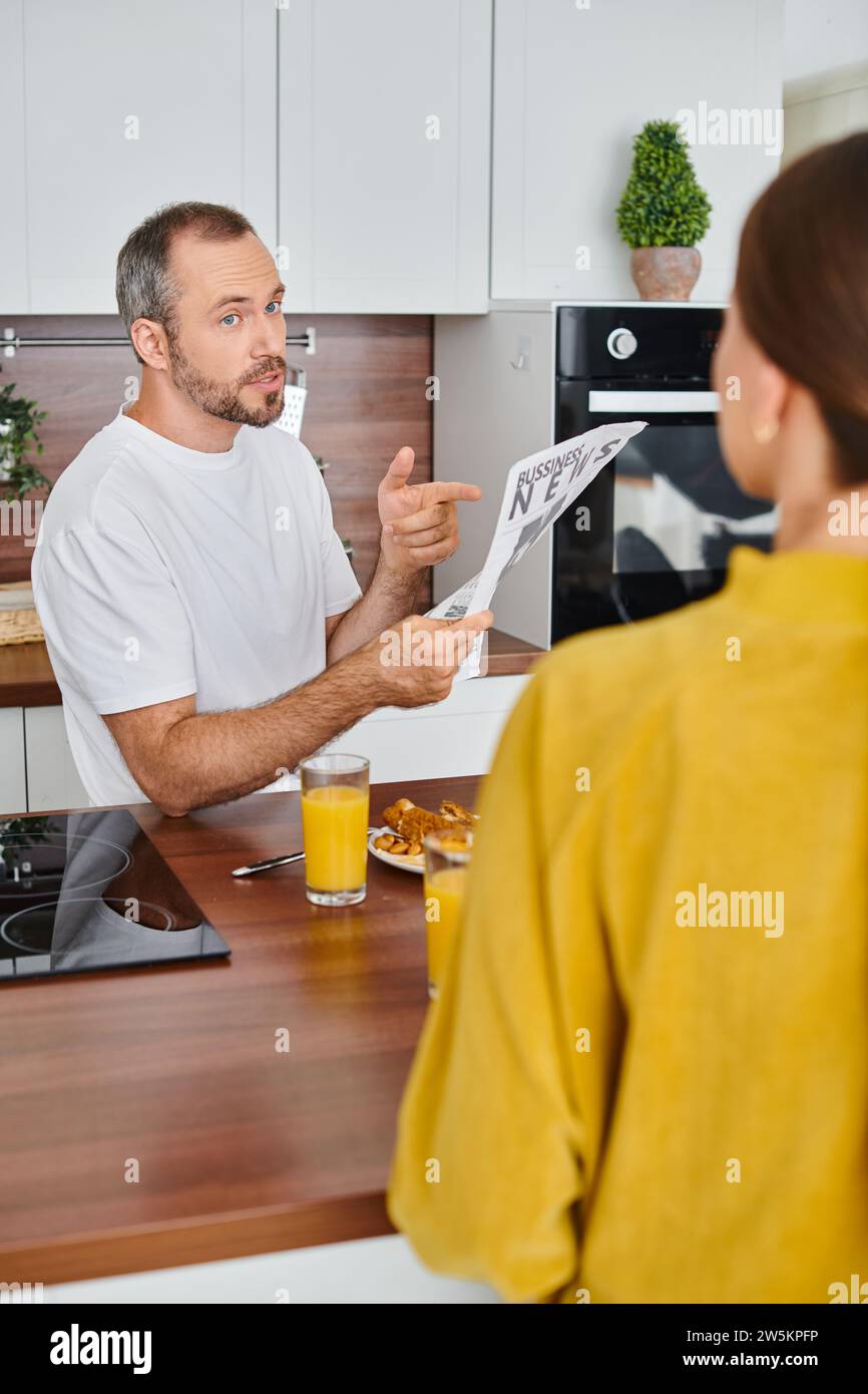 serious man pointing at newspaper near wife during breakfast in kitchen ...