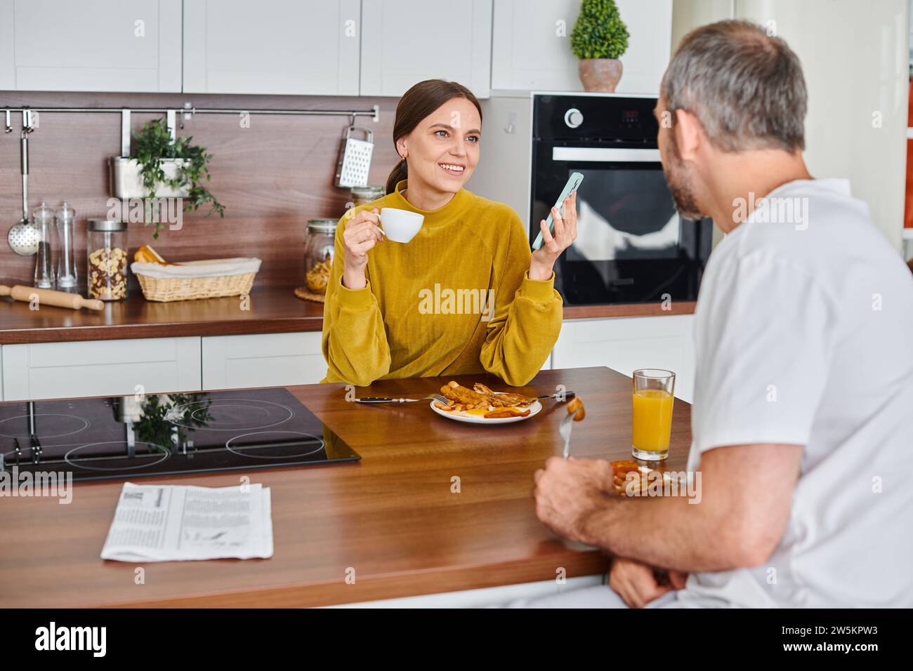 cheerful woman with coffee and smartphone talking to husband during breakfast, child-free ...