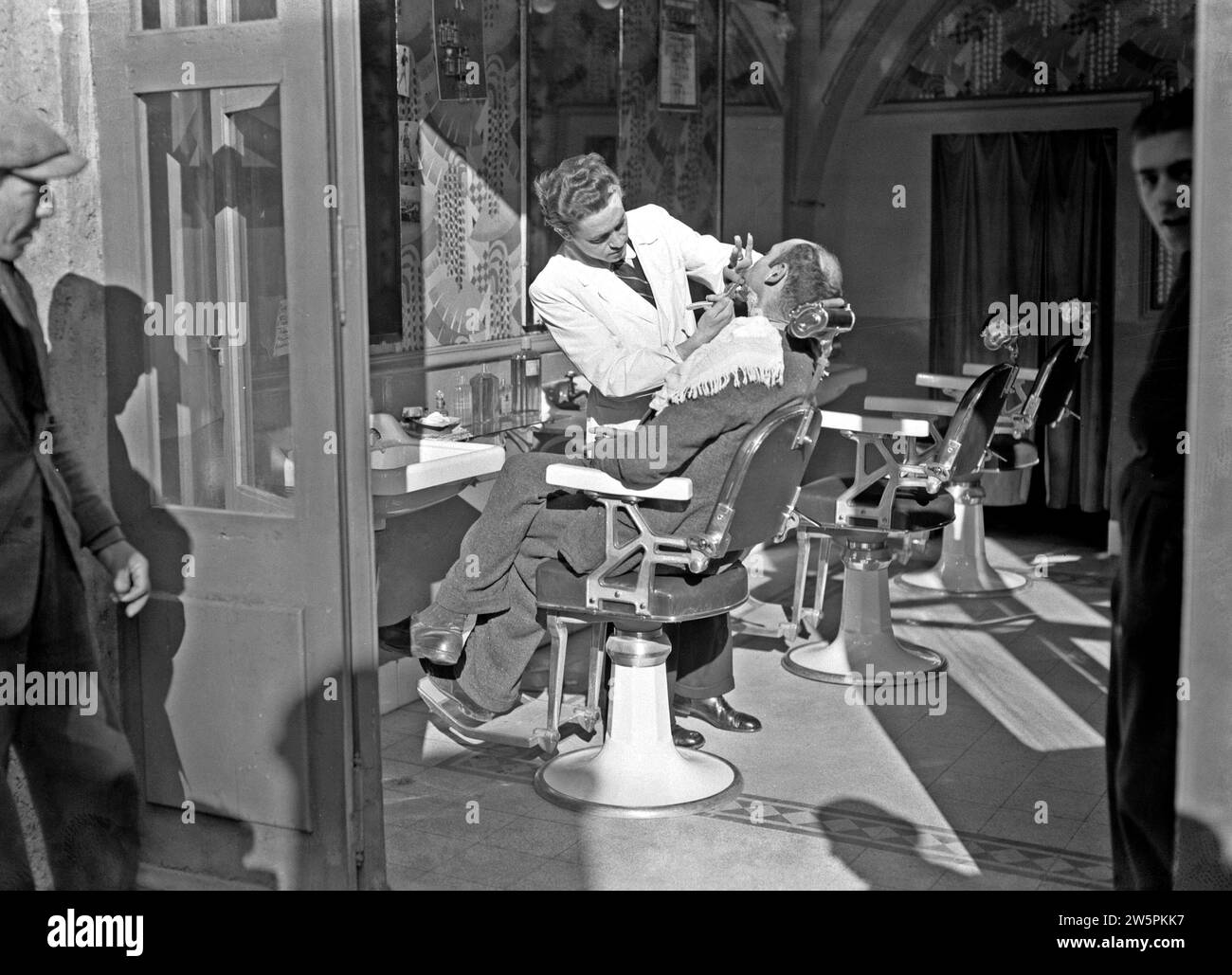 Man is shaved by a barber while two men watch in the doorway ca ...