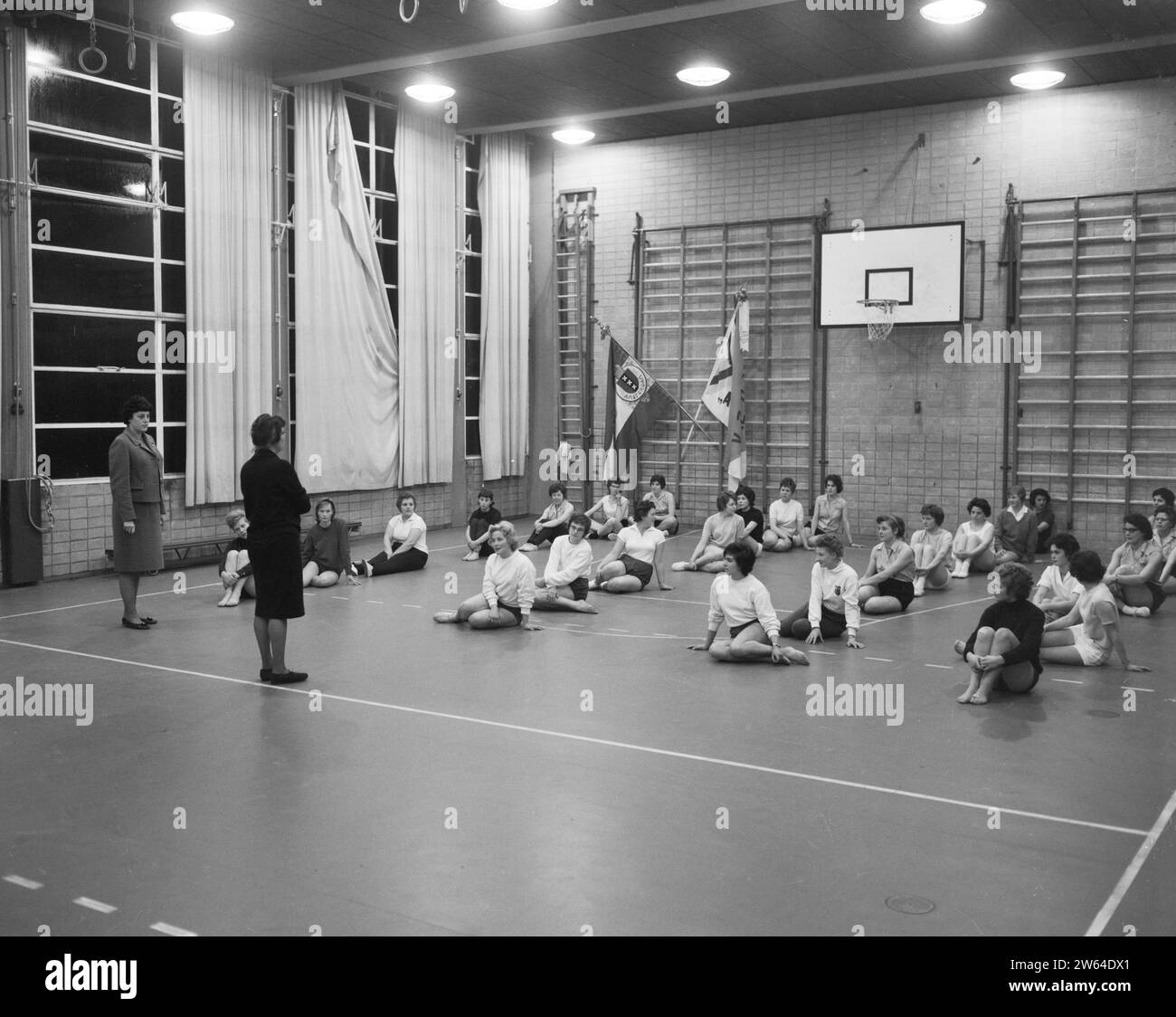 Adult women sitting in rows on the floor of a gymnasium, listening to a ...