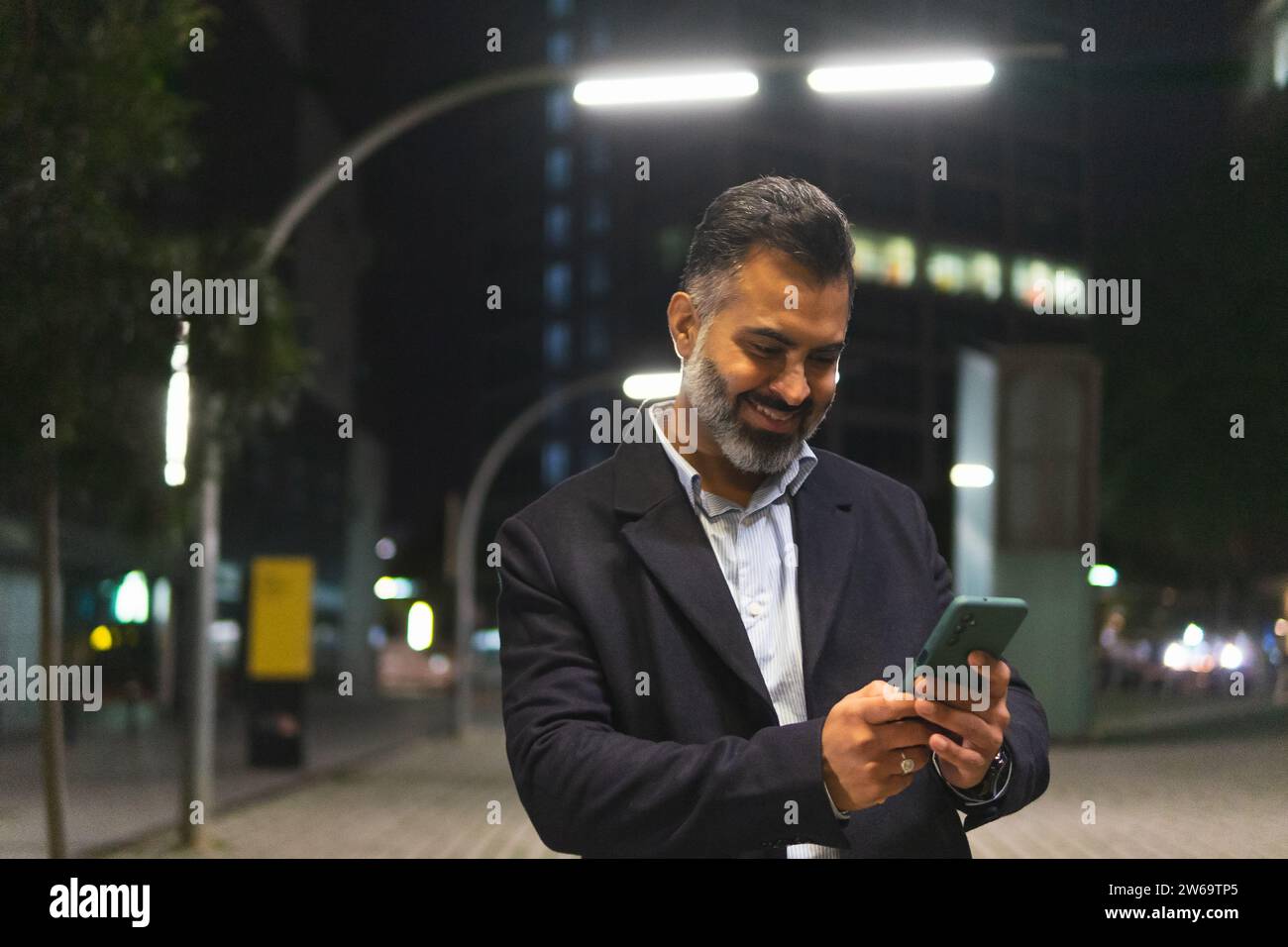 Smiling bearded indian man in suit standing on street with streetlights ...