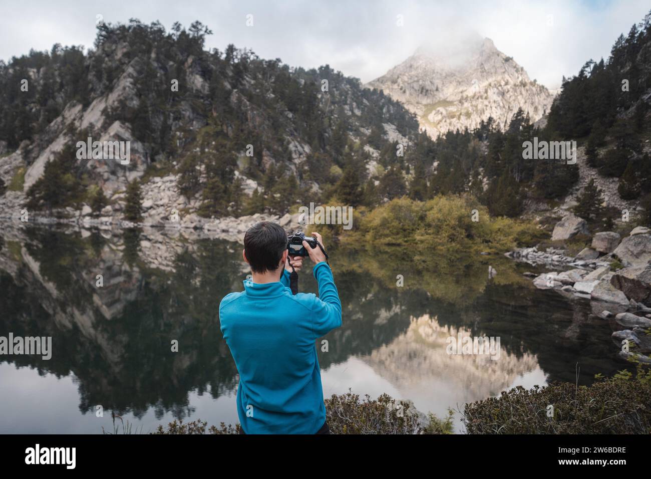Back view of unrecognizable male tourist standing on lakeshore photographing mountains through ...