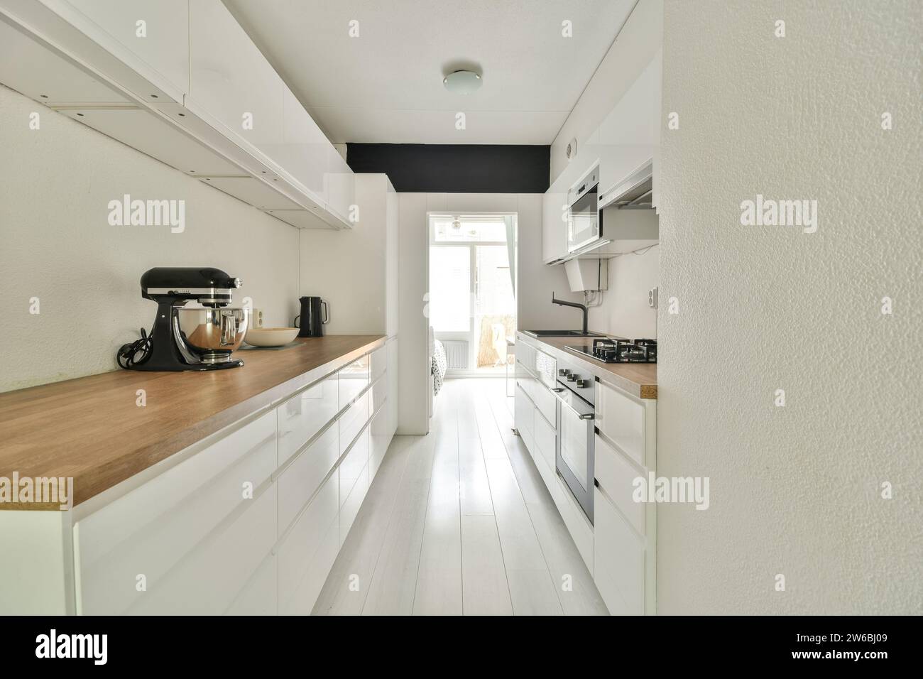 A bright, narrow modern kitchen featuring white cabinetry, wooden ...