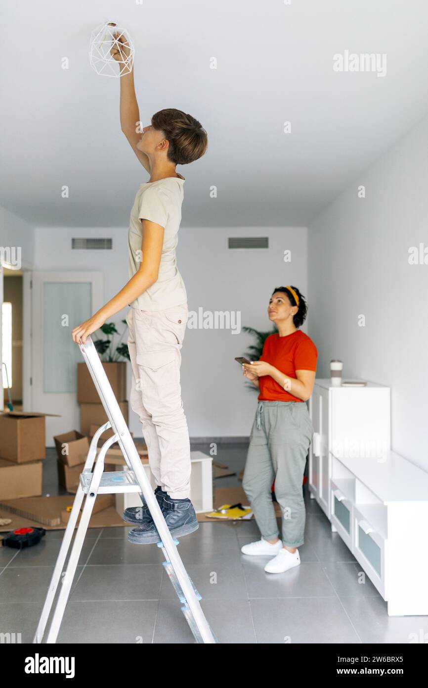 Side view of teen in gray casual clothes standing on ladder to fix lamp ...