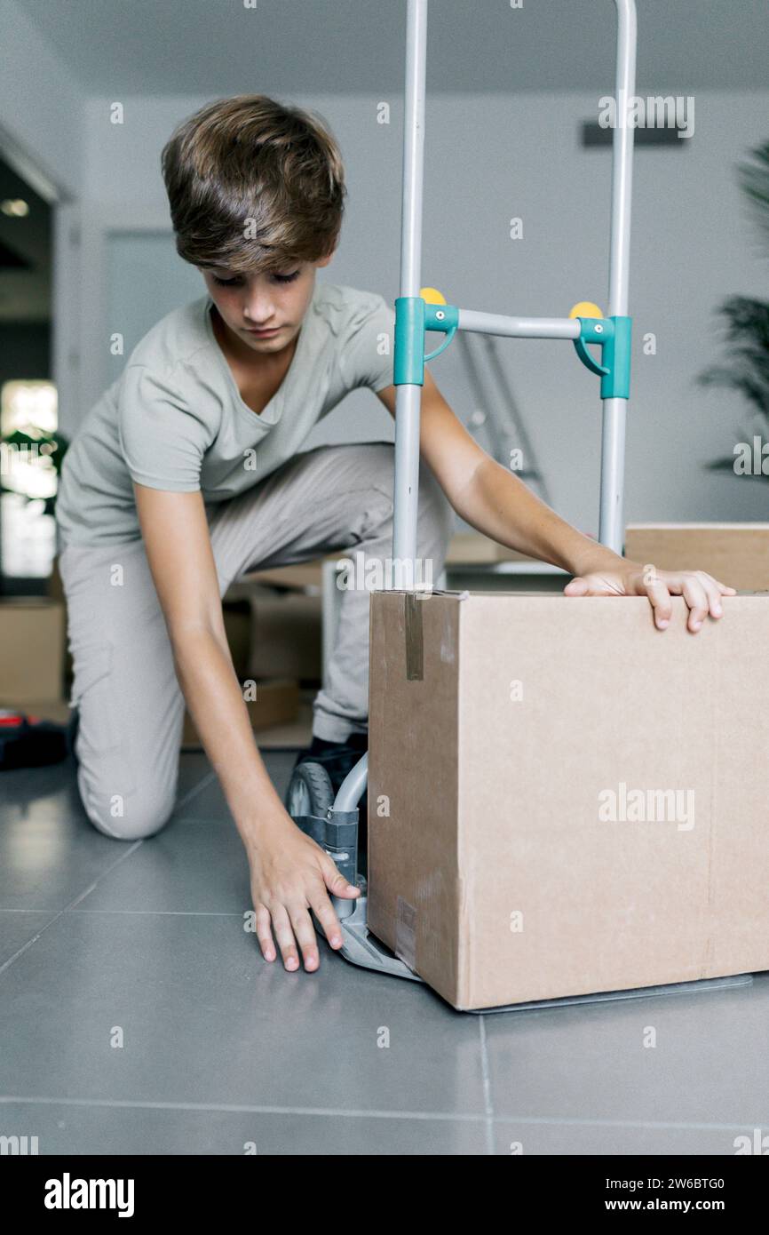 Focused brown haired teen in casual outfit kneeling and packing carton ...