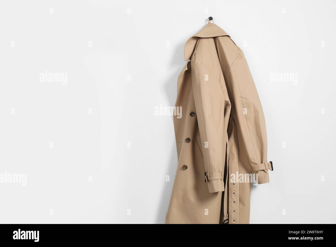 Beige trench coat hanging on hook rack on white wall, space for text ...