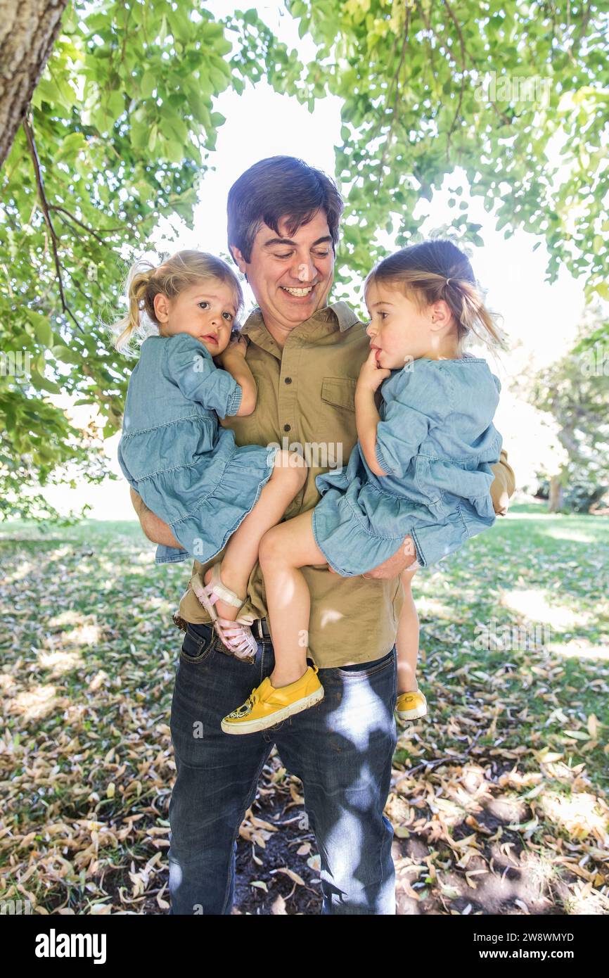 Father holding twin girls wearing matching dresses underneath tree ...