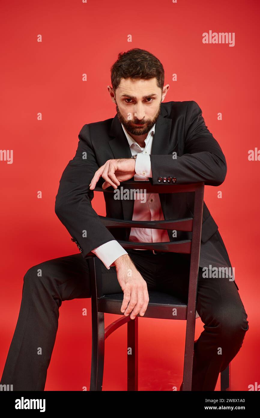 serious sharp-dressed man sitting on chair and looking at camera on red ...