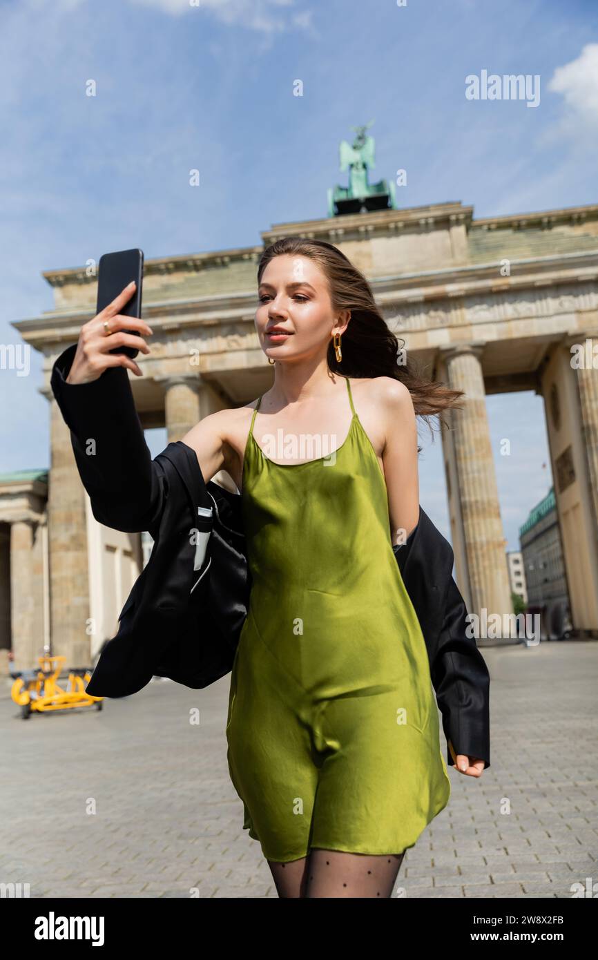 Fashionable woman in jacket and silk dress taking selfie near ...