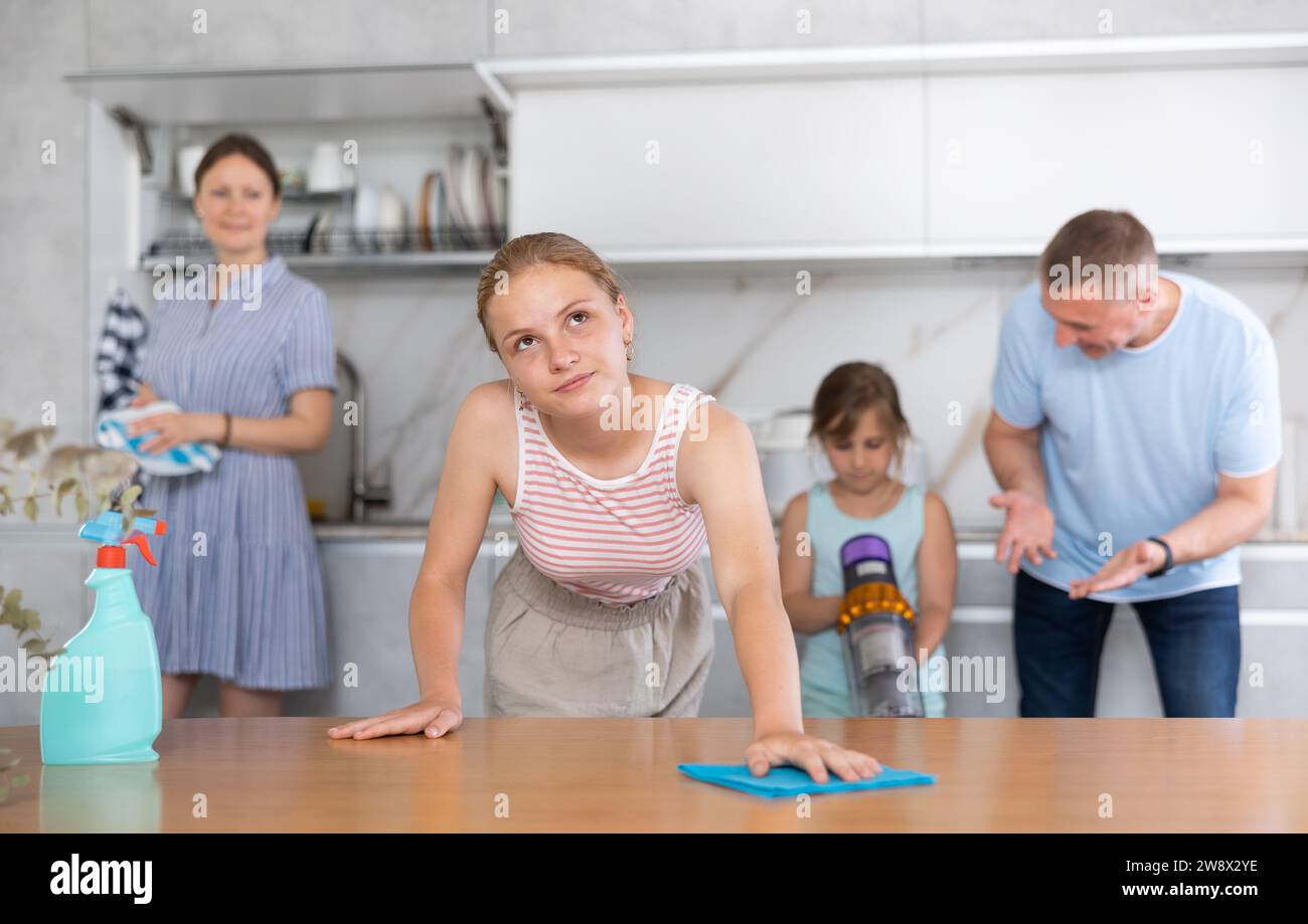 Teen girl cleans table with rag, family clean kitchen in background ...