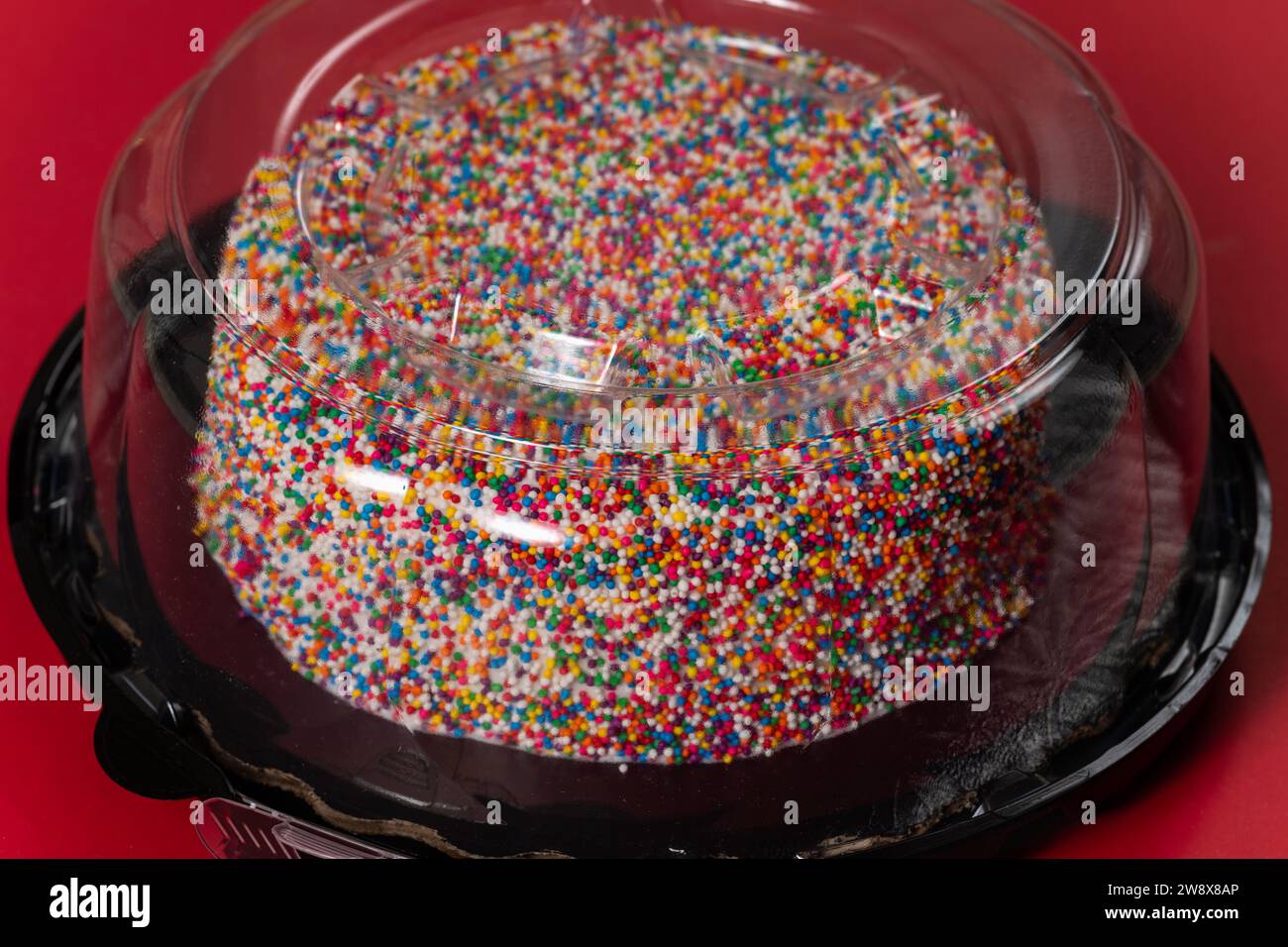 A cake with white icing encrusted in rainbow sprinkles in a plastic ...
