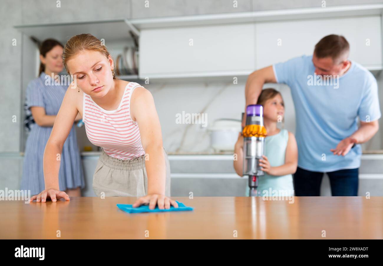 Teen girl cleans table with rag, family clean kitchen in background ...