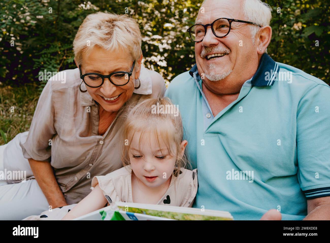 Happy grandparents sitting with granddaughter reading book at park ...