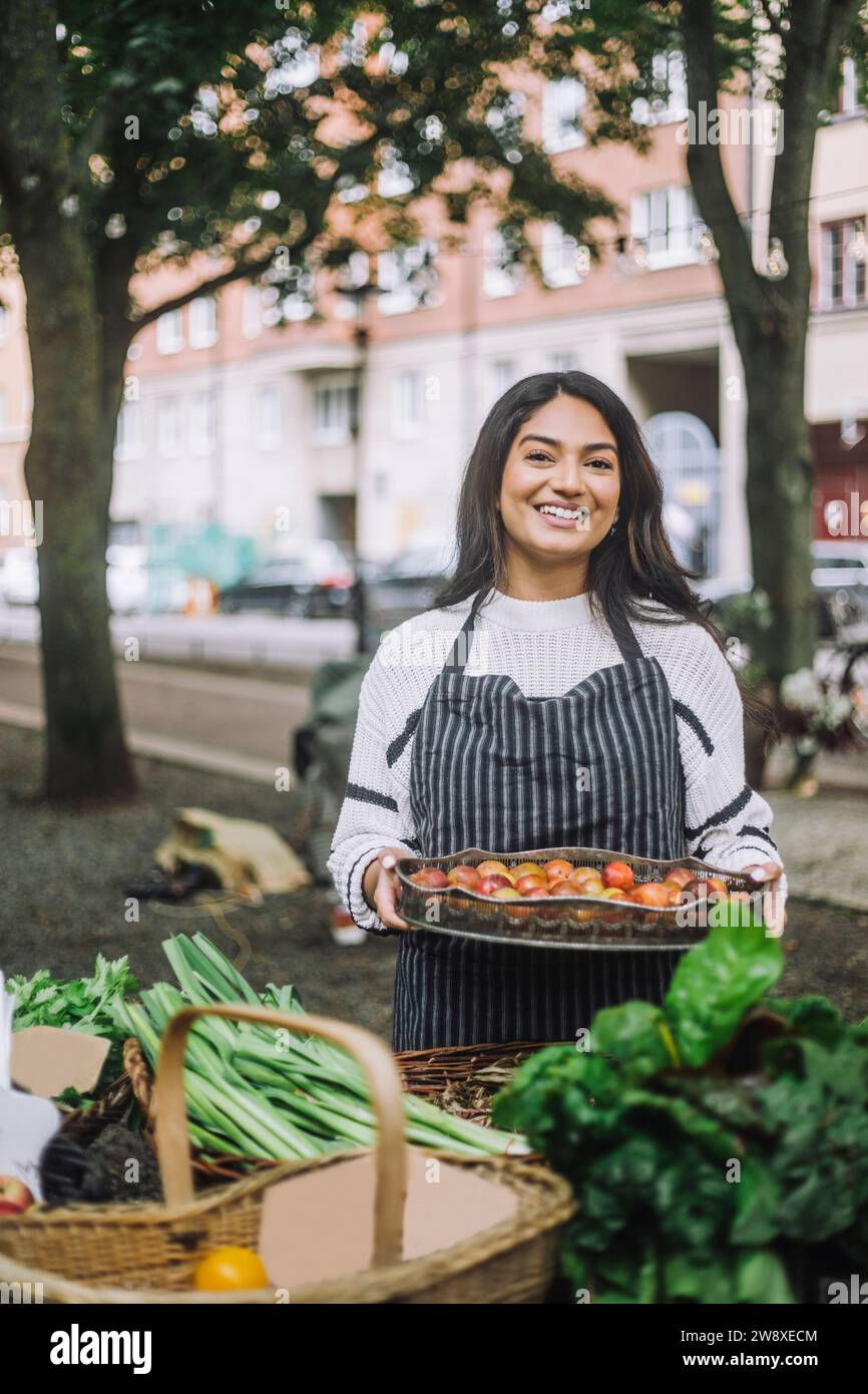 Portrait of young female vendor holding vegetable tray near stall at ...