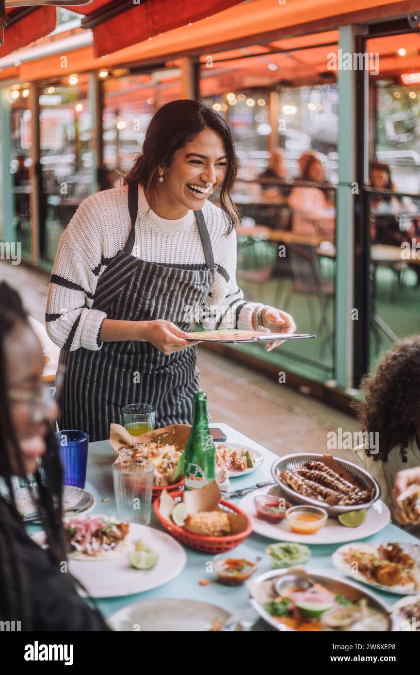 Happy waitress laughing while talking to customers near table at ...